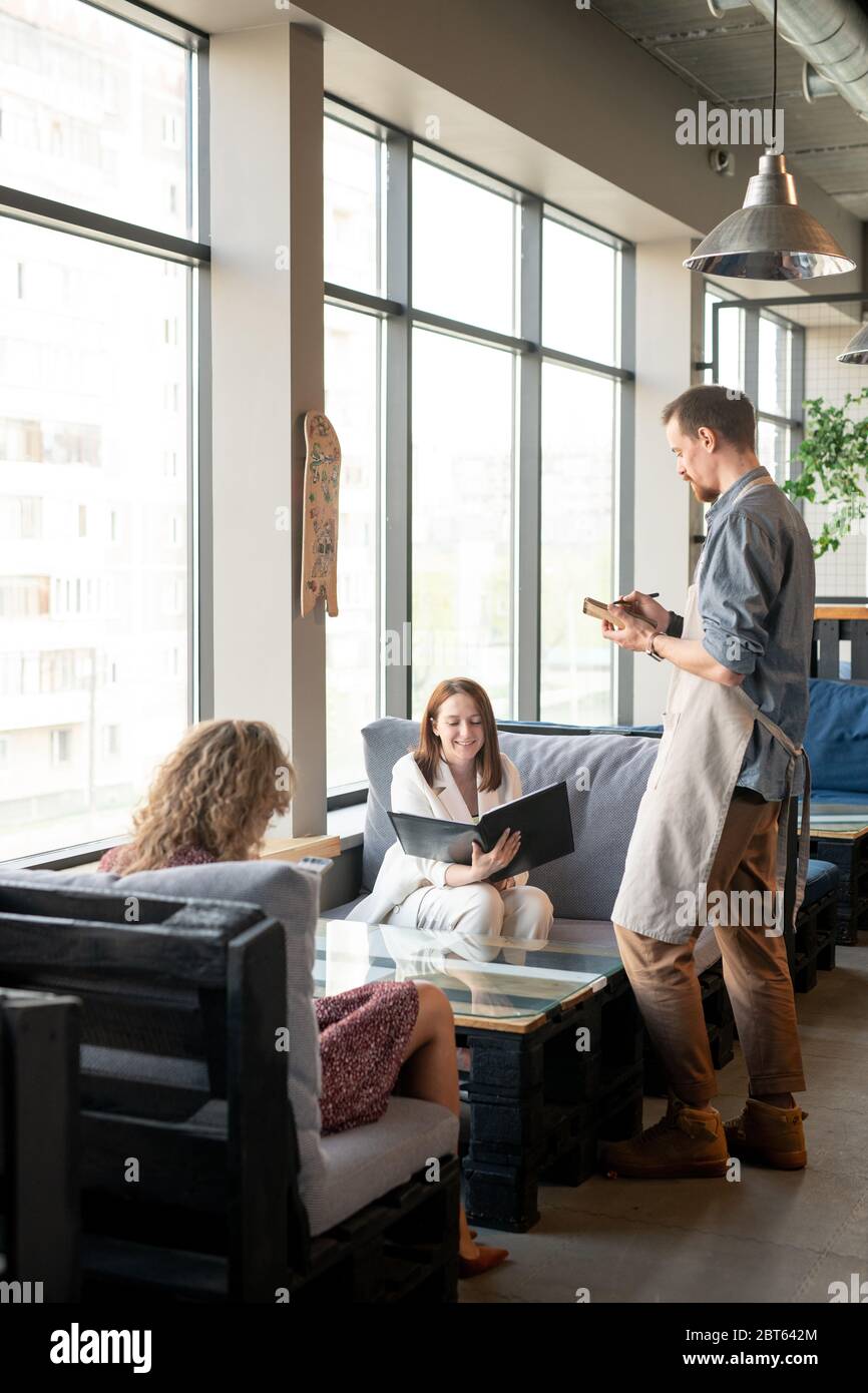 Two contemporary young women sitting on couches by table and choosing ...
