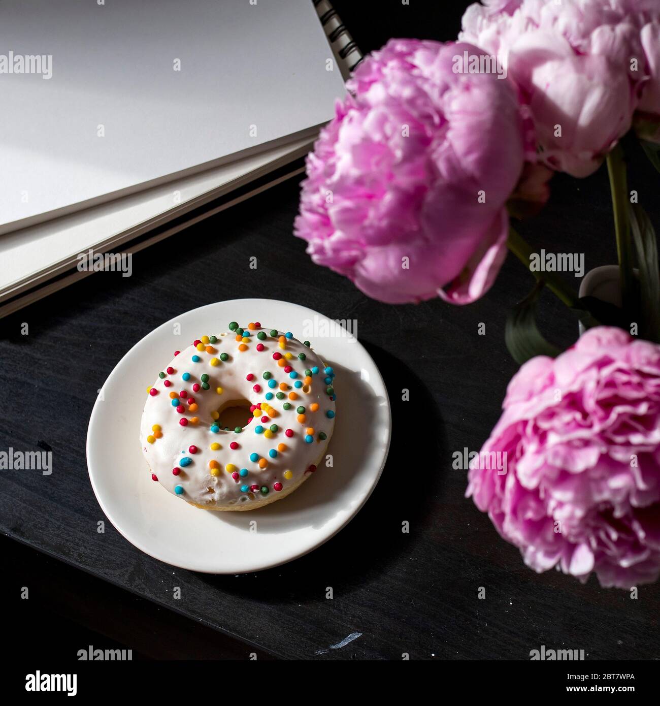 White donut with colored dusting on a saucer and a couple of peonies on ...