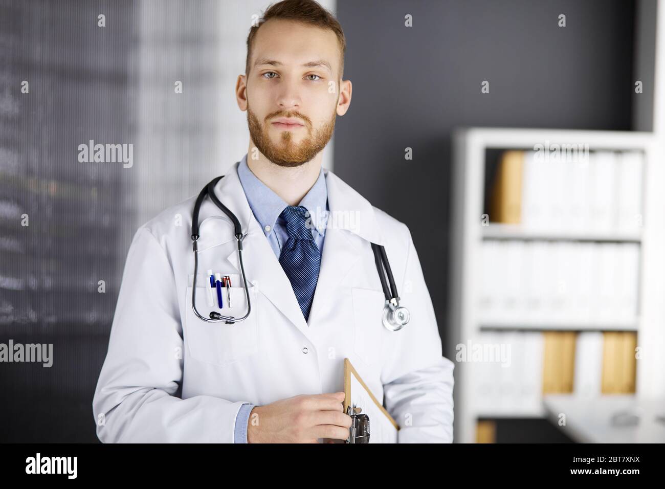 Red-bearded doctor standing straight in clinic near his working place ...