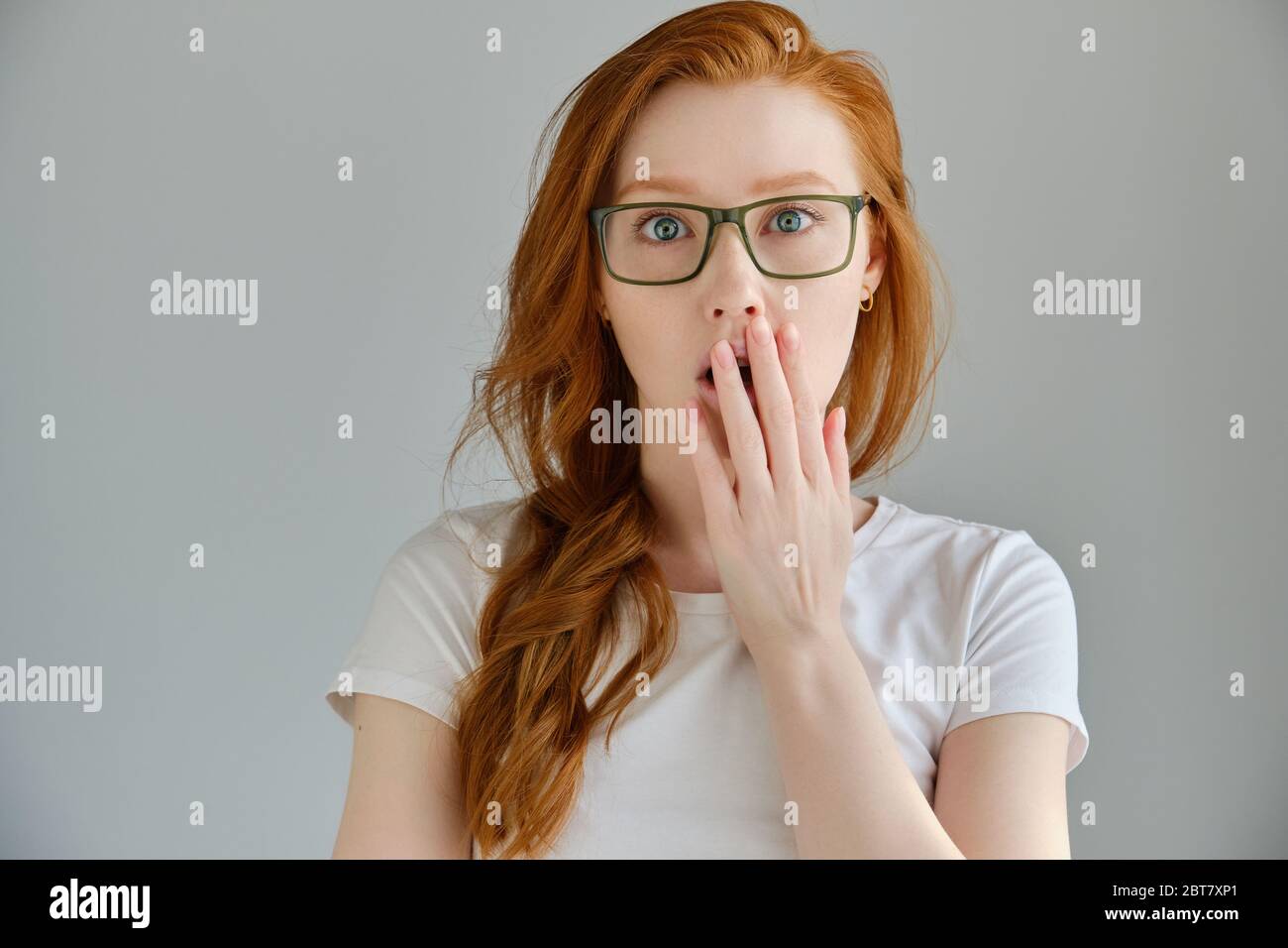 Redhead girl in a white T-shirt and glasses stands on a gray background ...