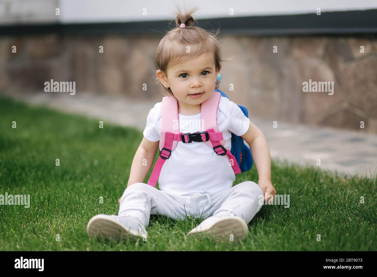 Adorable little girl sitting on the grass with backpack. Cute little girl outdoors Stock Photo ...