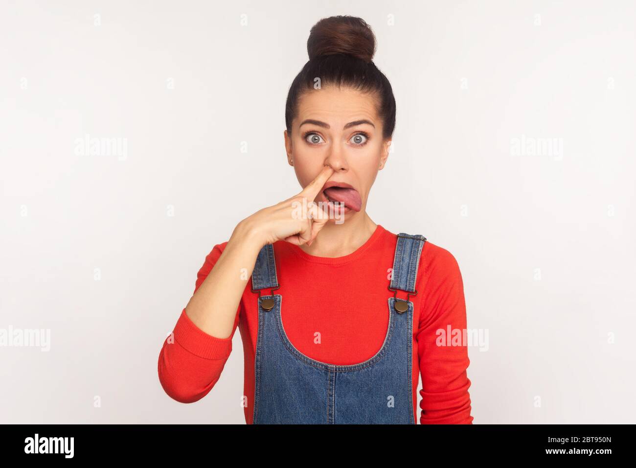 Portrait of funny comical girl with hair bun in denim overalls picking ...