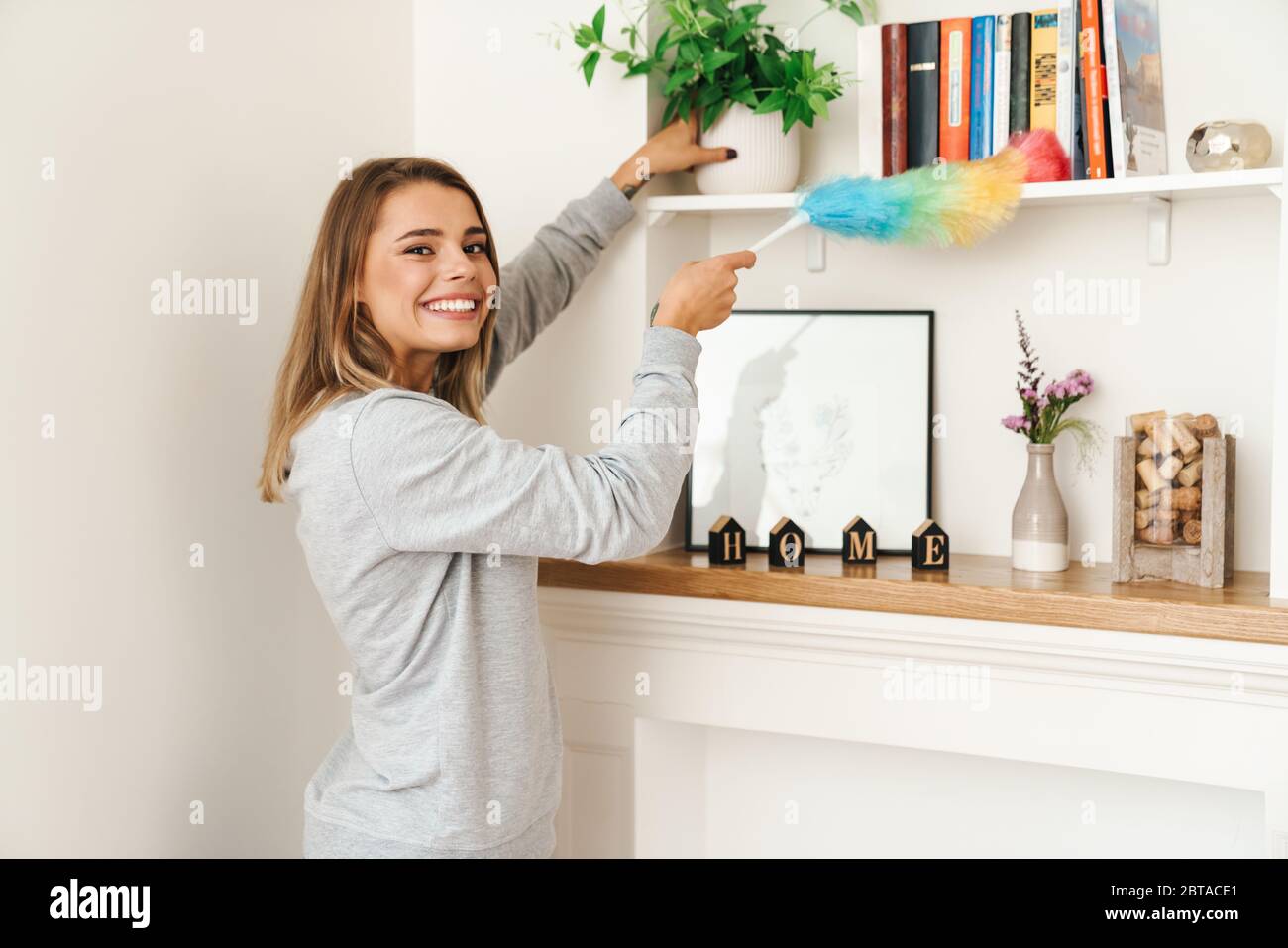 Photo of joyful young beautiful woman homemaker smiling and doing housework with duster at ...