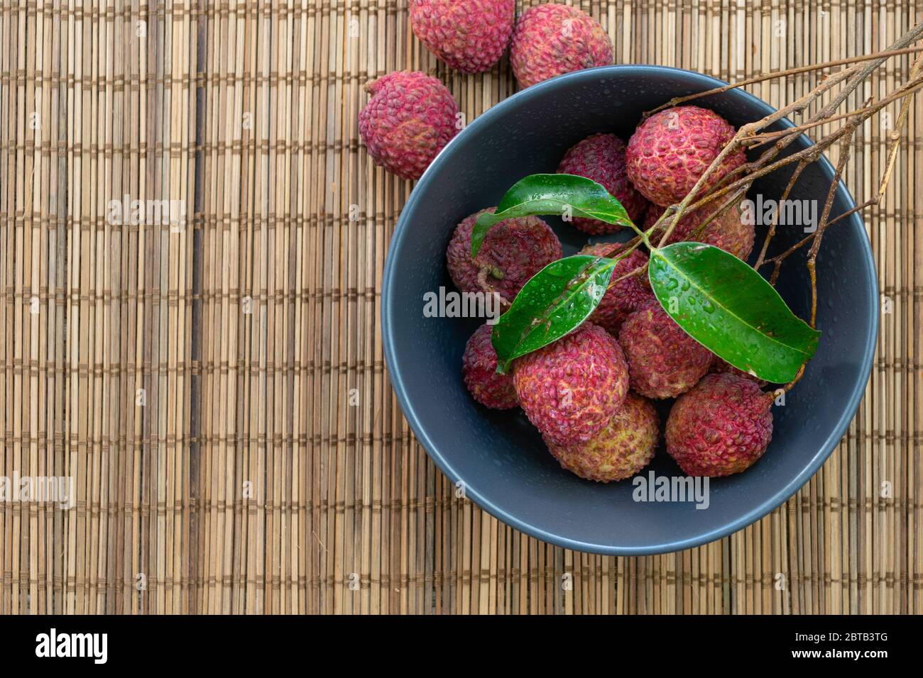 Lychee with water drop in black bowl over brown bamboo mat. Top view ...