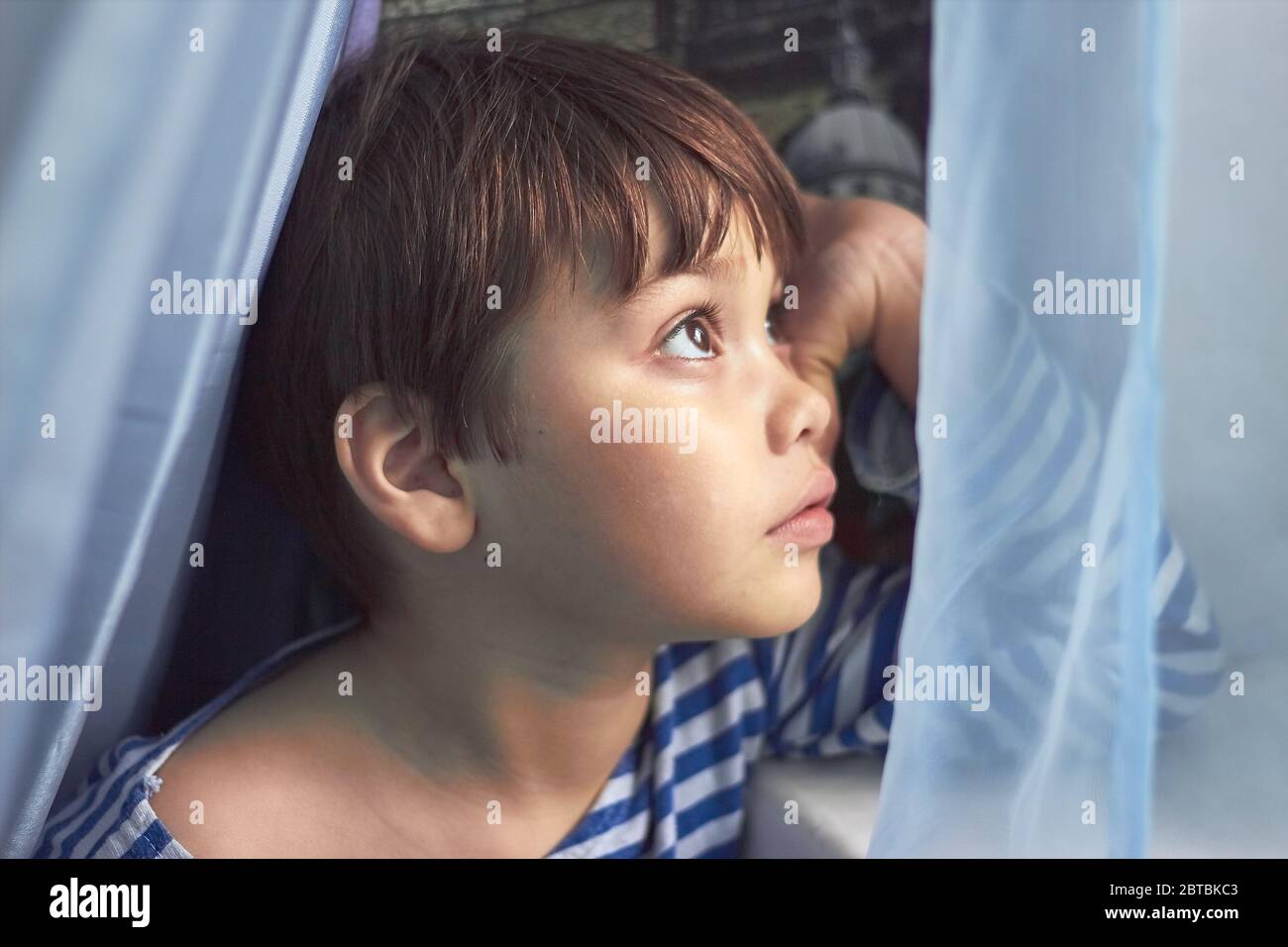 small beautiful boy on self isolation looks out the window with hope ...