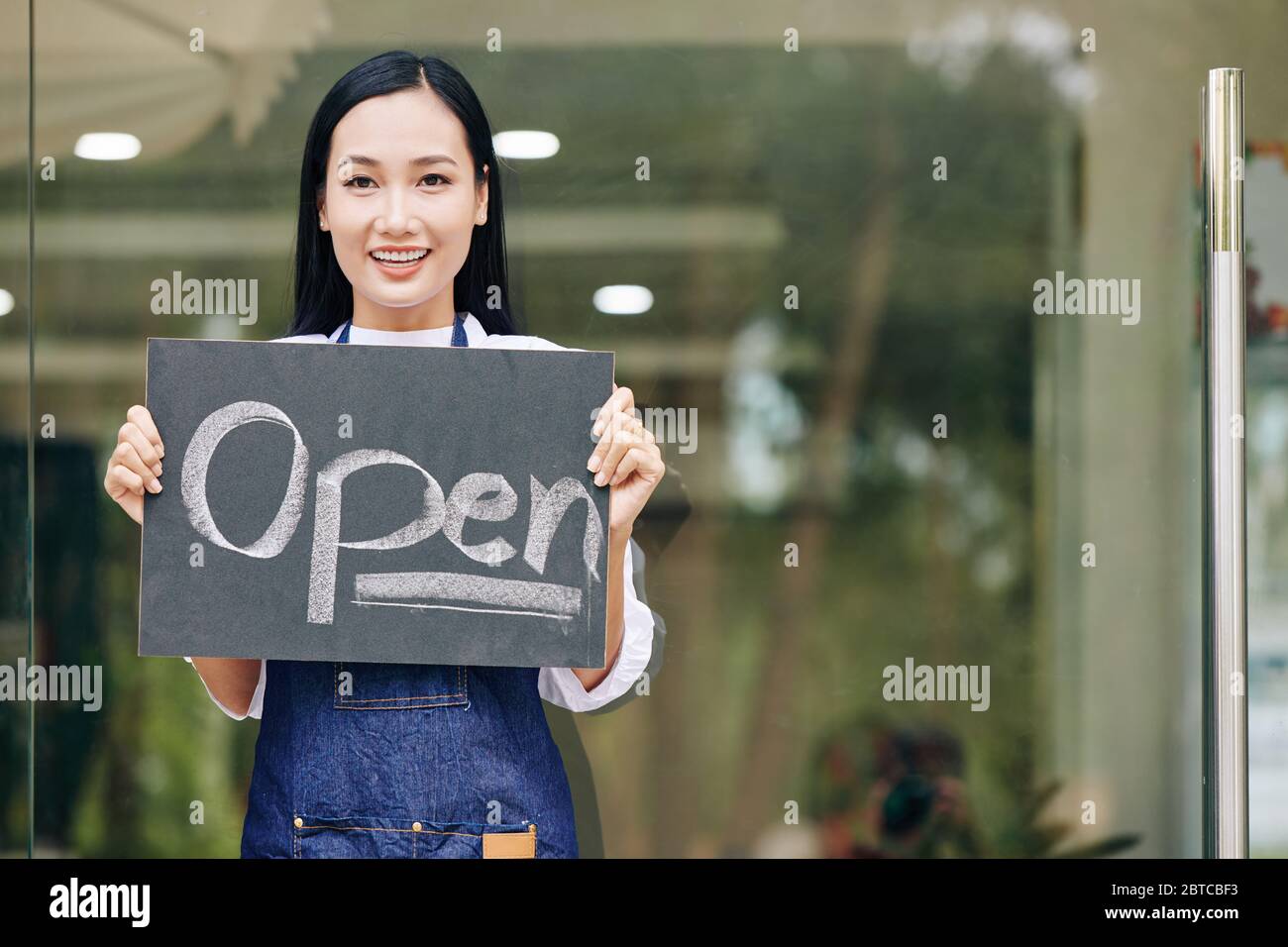 Portrait of cheerful young female cafe owner showing open sign written ...