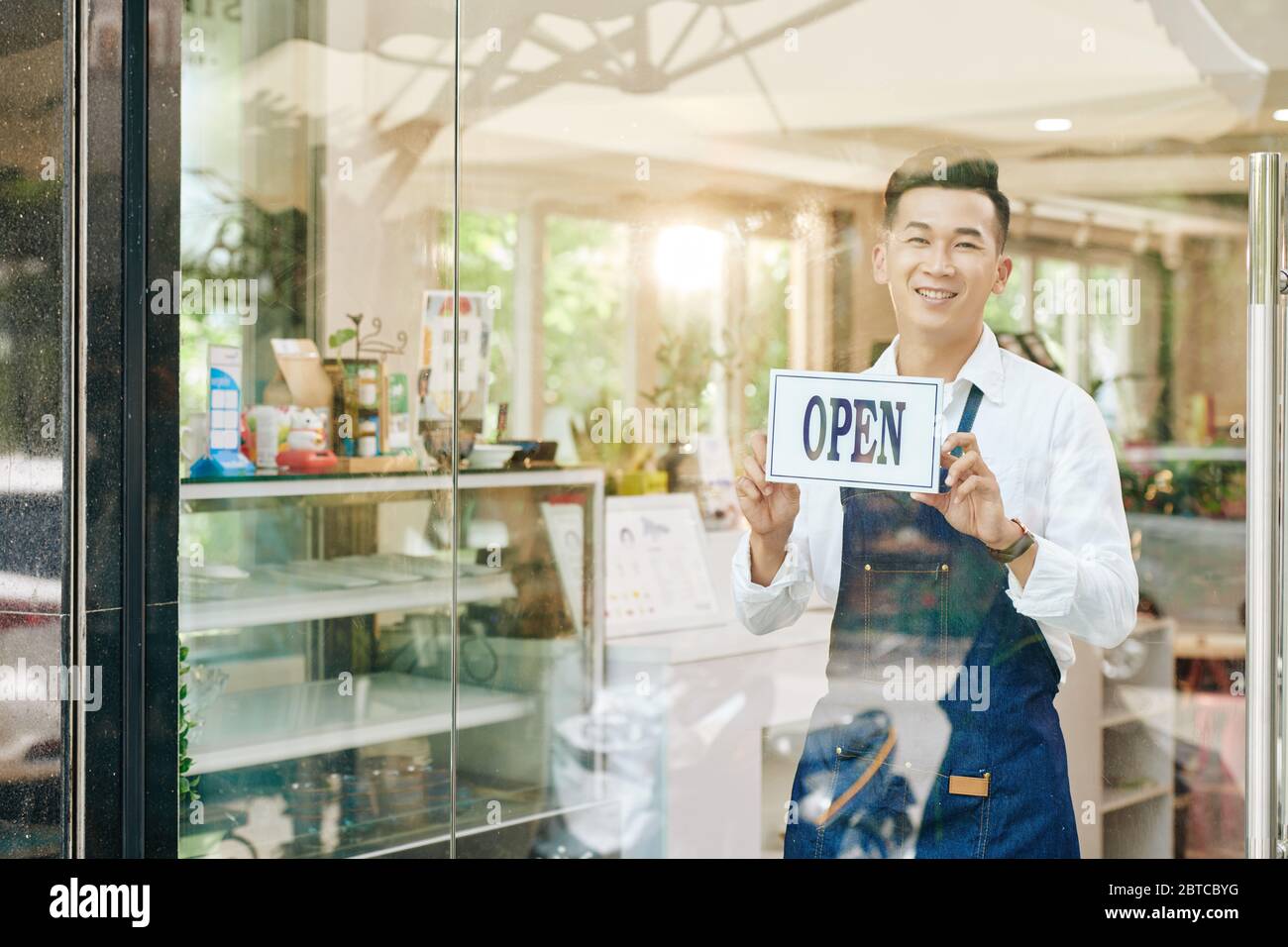 Portrait of cheerful young Vietnamese waiter hanging open sign on glass ...