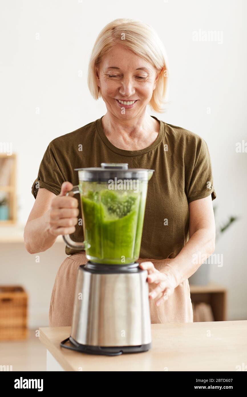 Happy mature woman making juice from fresh vegetable in blender and ...