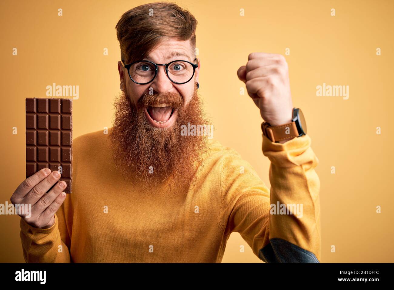 Irish redhead man with beard eating a sweet chocolate bar over yellow ...