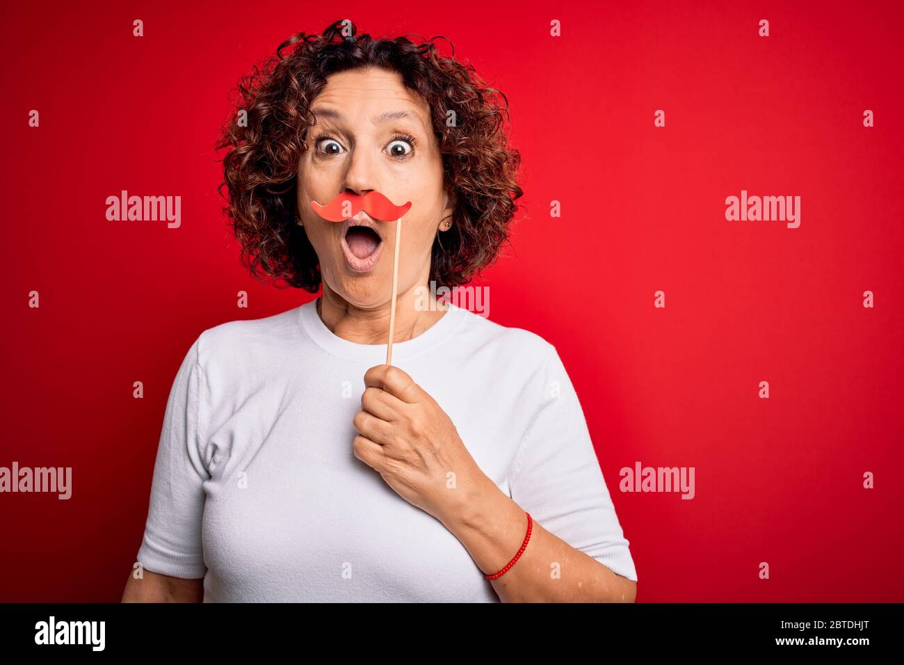 Middle age curly hair woman holding funny mustache over isolated red ...