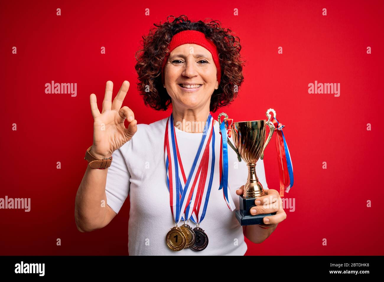Middle age curly woman winning medals holding trophy over isolated red ...
