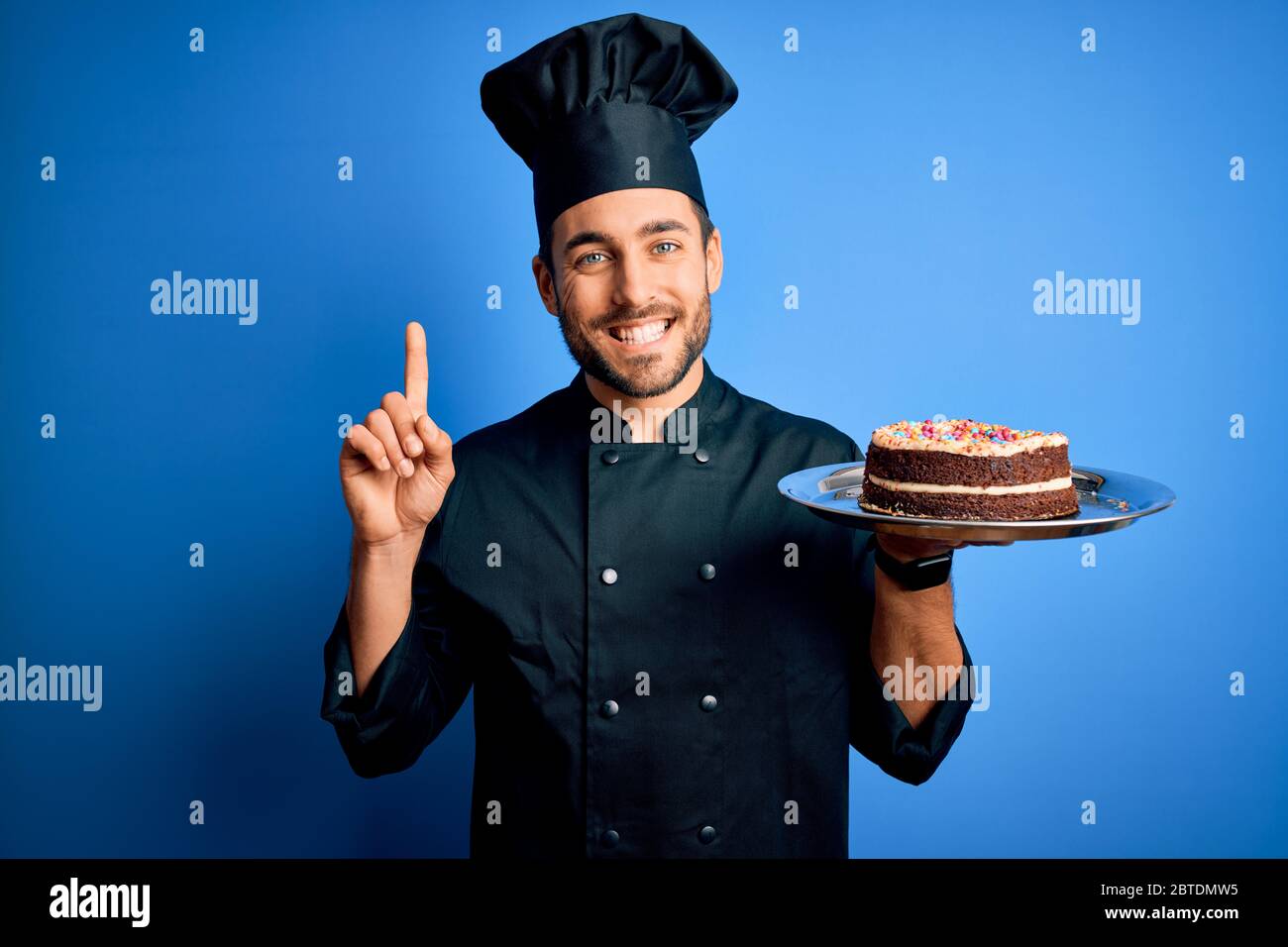 Young handsome cooker man with beard wearing uniform and hat holding ...