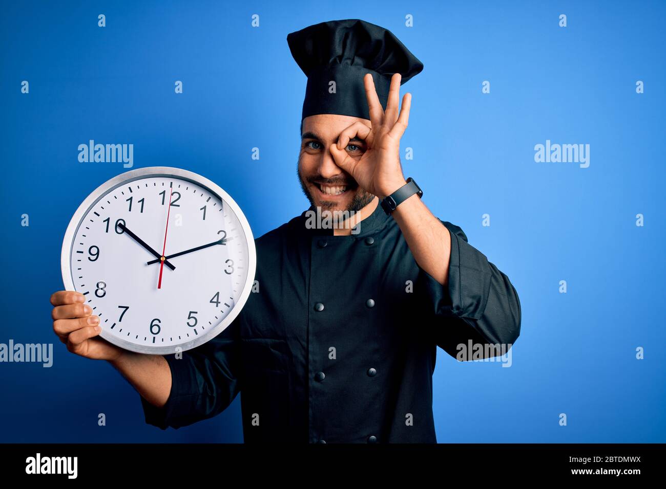 Young handsome cooker man with beard wearing uniform holding clock ...