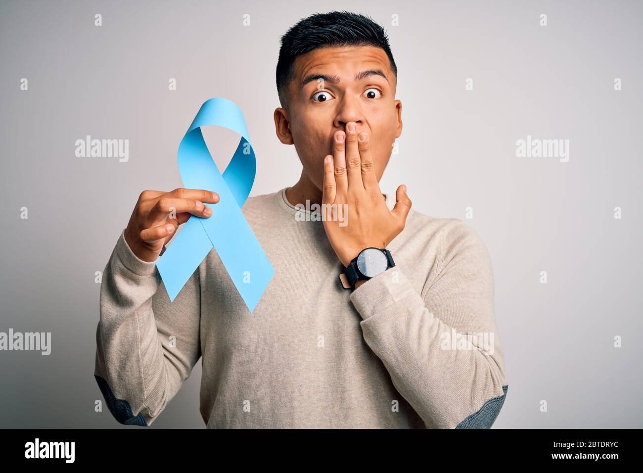 Young handsome latin man holding blue cancer ribbon over isolated white ...