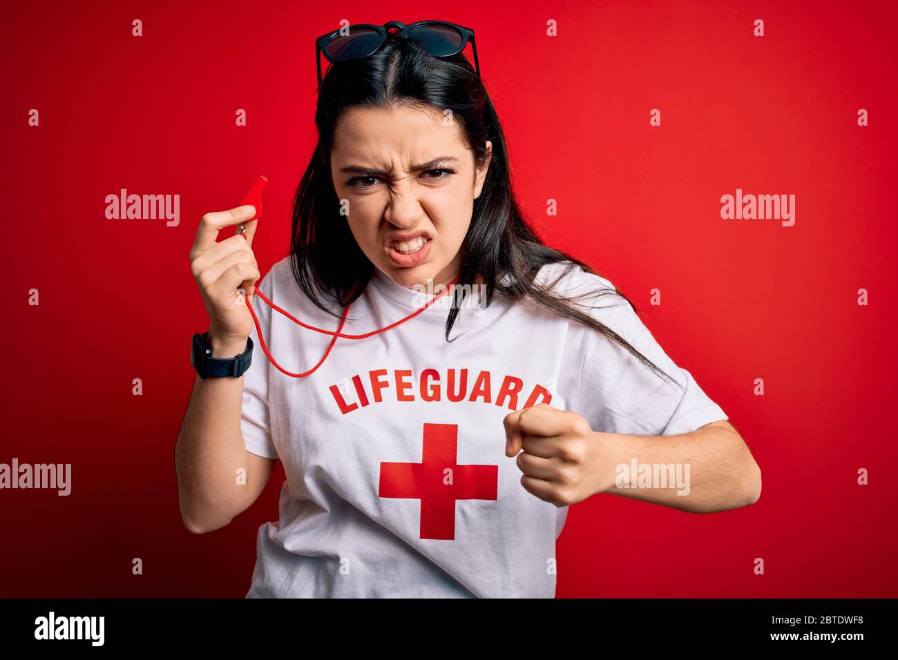 Young lifeguard woman wearing guard equipement holding whistle over red ...
