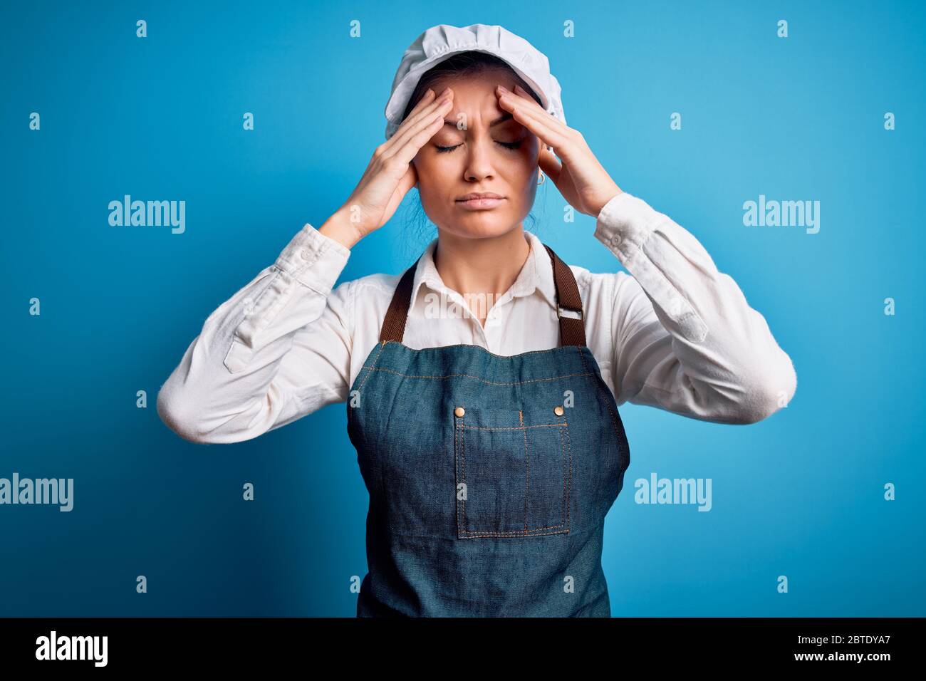 Young beautiful baker woman with blue eyes wearing apron and cap over ...