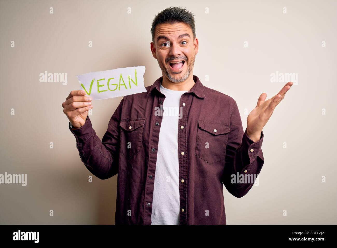 Young handsome veggie man holding paper with vegan message over white ...