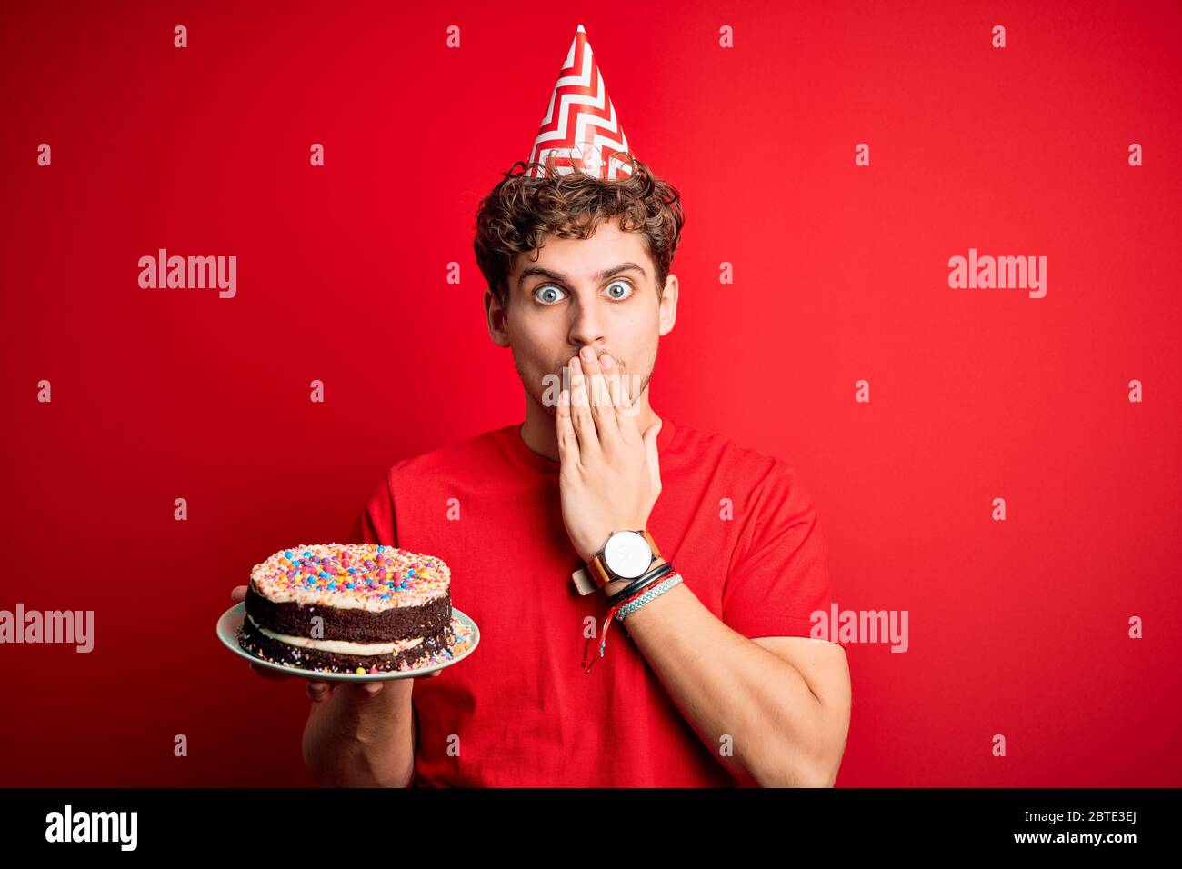 Young blond man with curly hair wearing birthday hat holding cake over ...