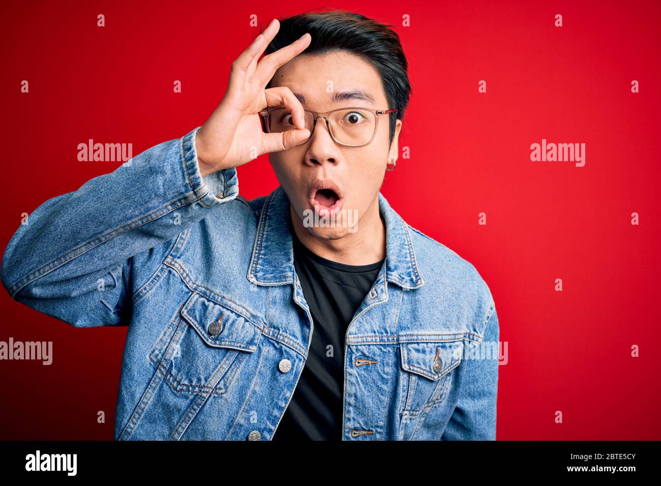 Young handsome chinese man wearing denim jacket and glasses over red ...