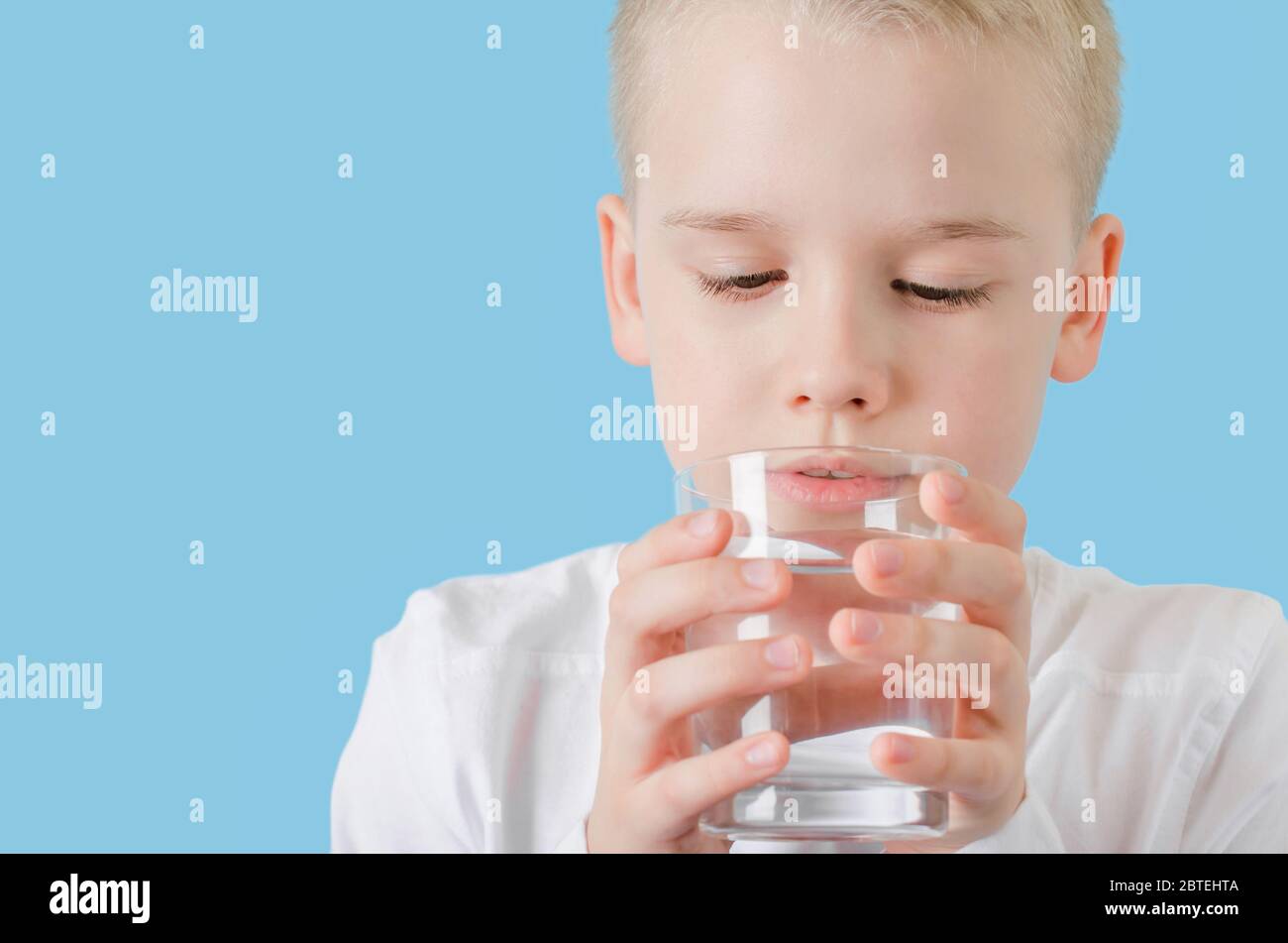 Cute child drinking clean water from glass. Blond little boy with water glass isolated on blue ...
