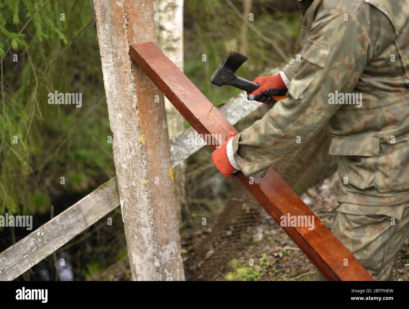A worker fixing a fence with a new wooden plank and an axe in his hands ...