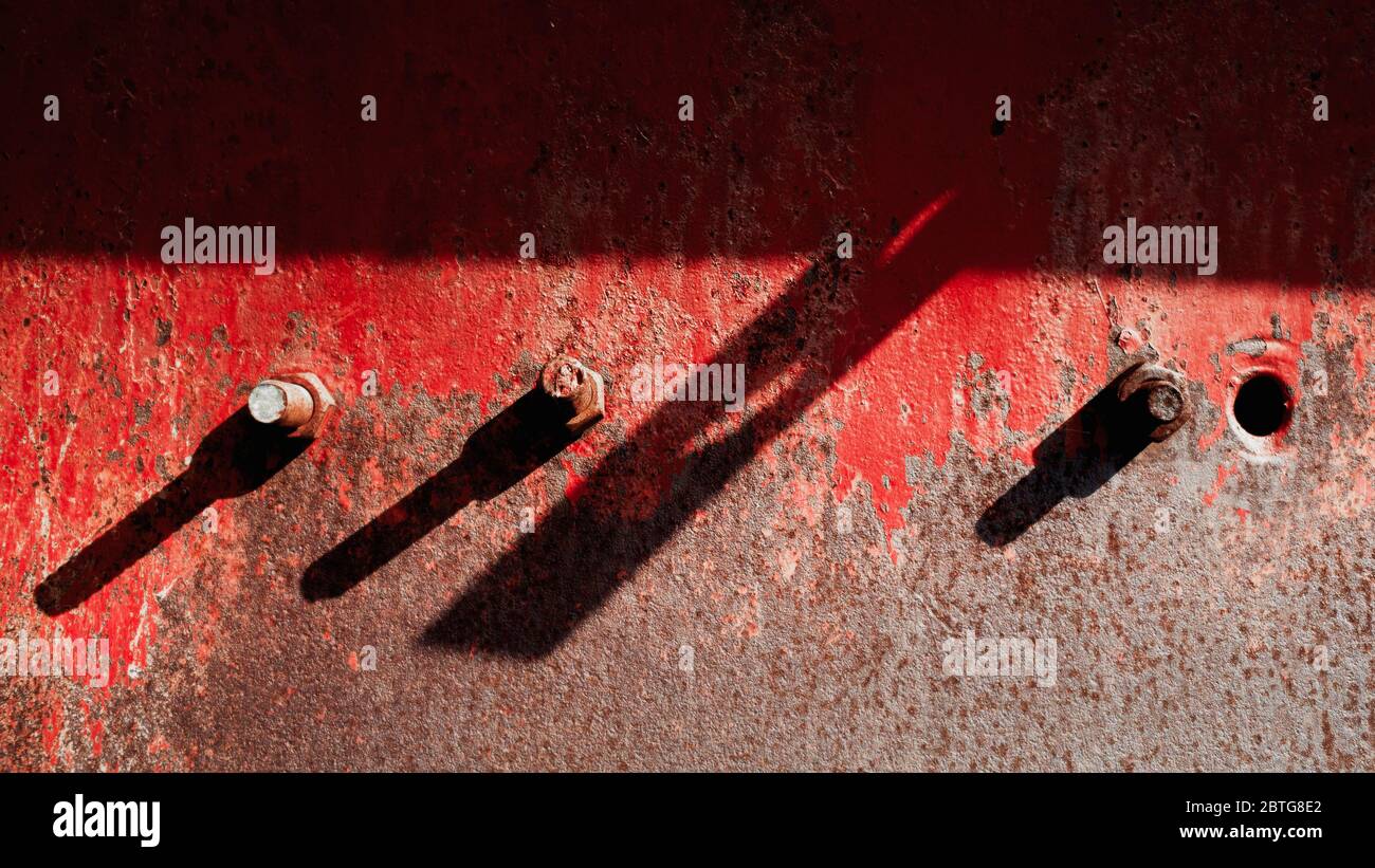 Red painted rusted metal and heavy bolts in sunlight with long shadows ...