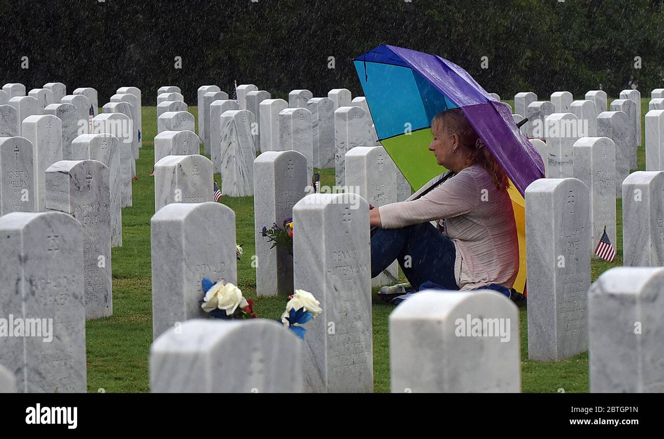 A woman holding an umbrella sits by a grave in the rain with a bouquet ...