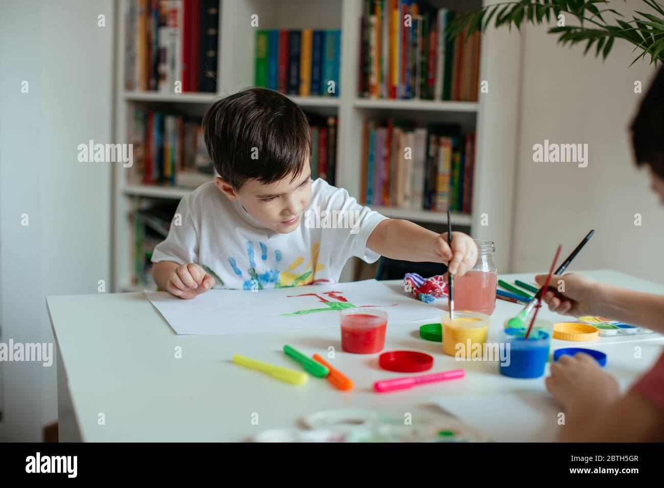 Left handed little boy reaching for paint with paintbrush in his hand ...