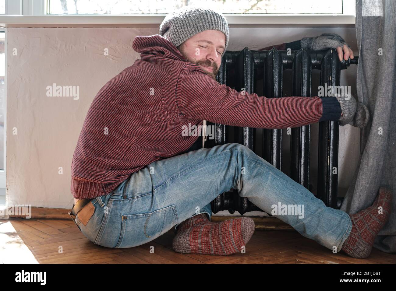 Man in warm clothes hugs a hot radiator sitting on the floor, he is ...