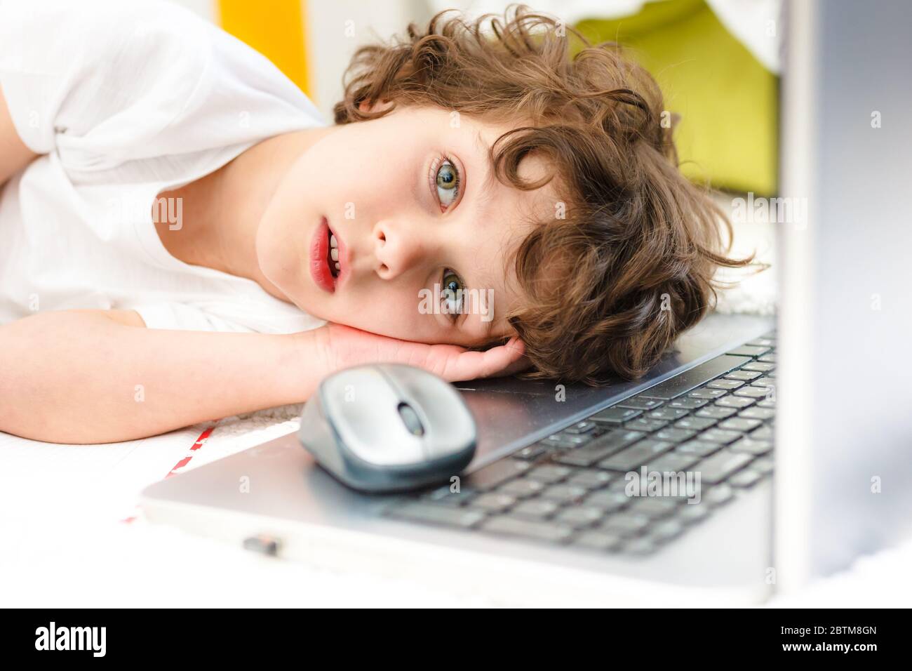Curly boy is engaged at computer. Tired child lies covering his face ...