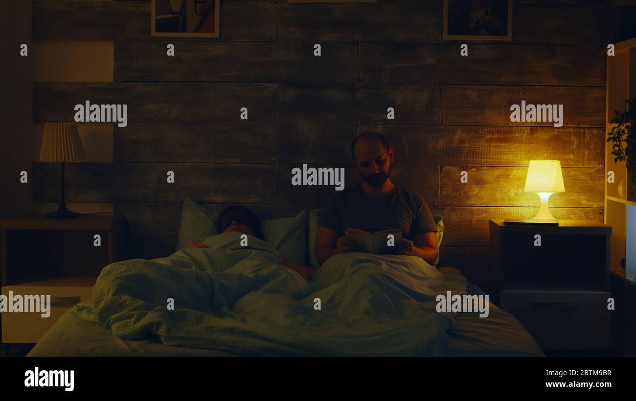 Young man reading a book while his wife is turning the lights off to ...