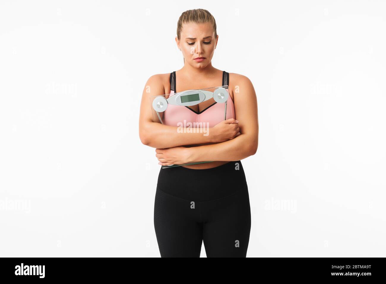Young upset woman with excess weight in sporty top holding scales in ...