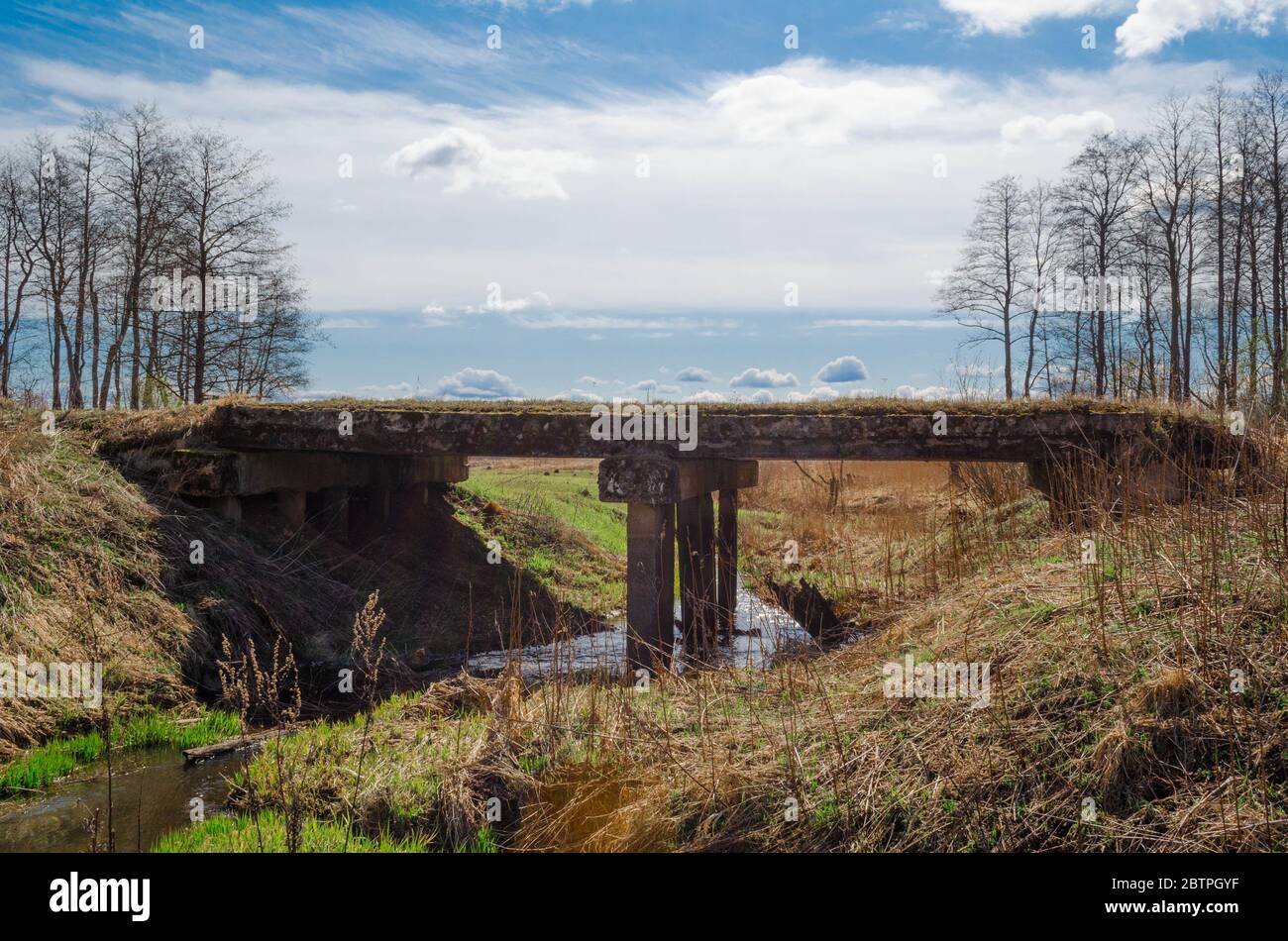 An old dilapidated bridge connects the road over a stream. Blue sky ...