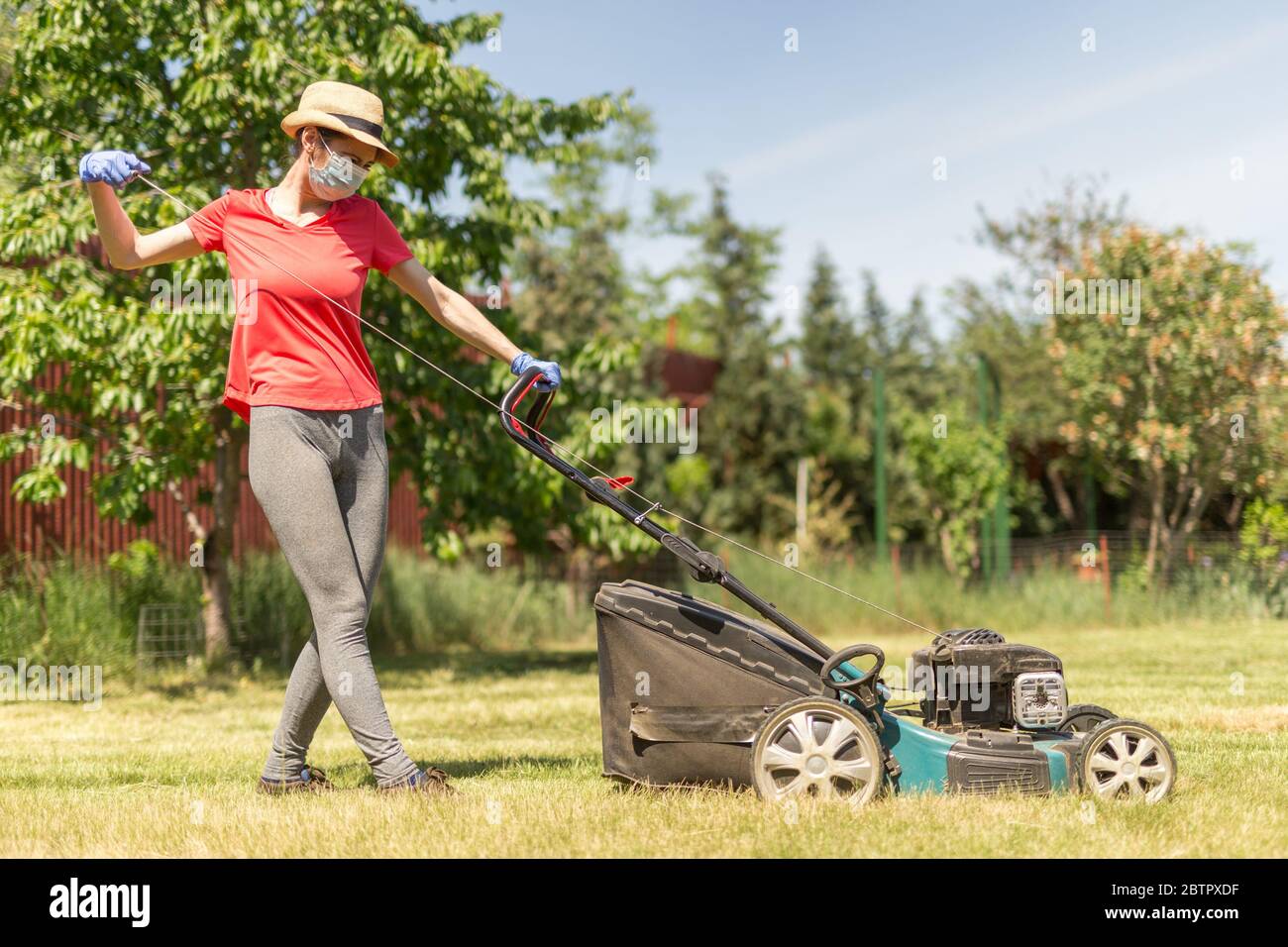 A woman in her backyard mowing grass with a lawn mower on a sunny day at home wearing a surgical ...