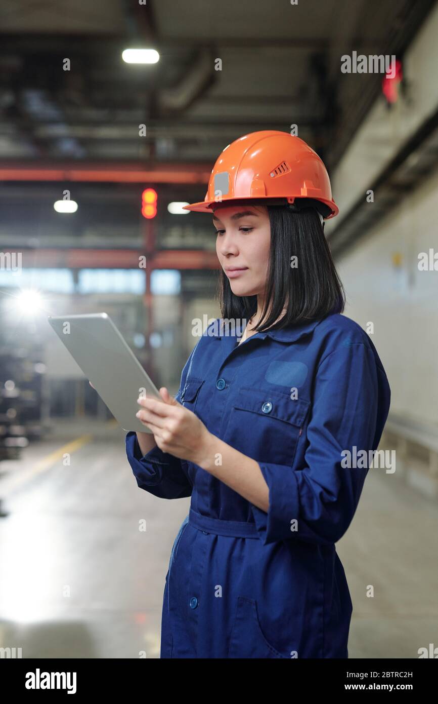 Serious young Asian female engineer in overall analyzing industrial ...