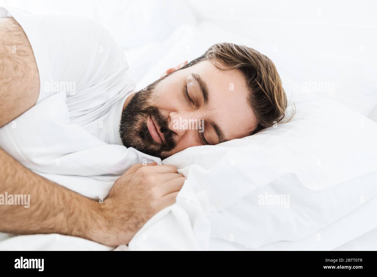 Image of young unshaven caucasian man sleeping alone in bed with white ...