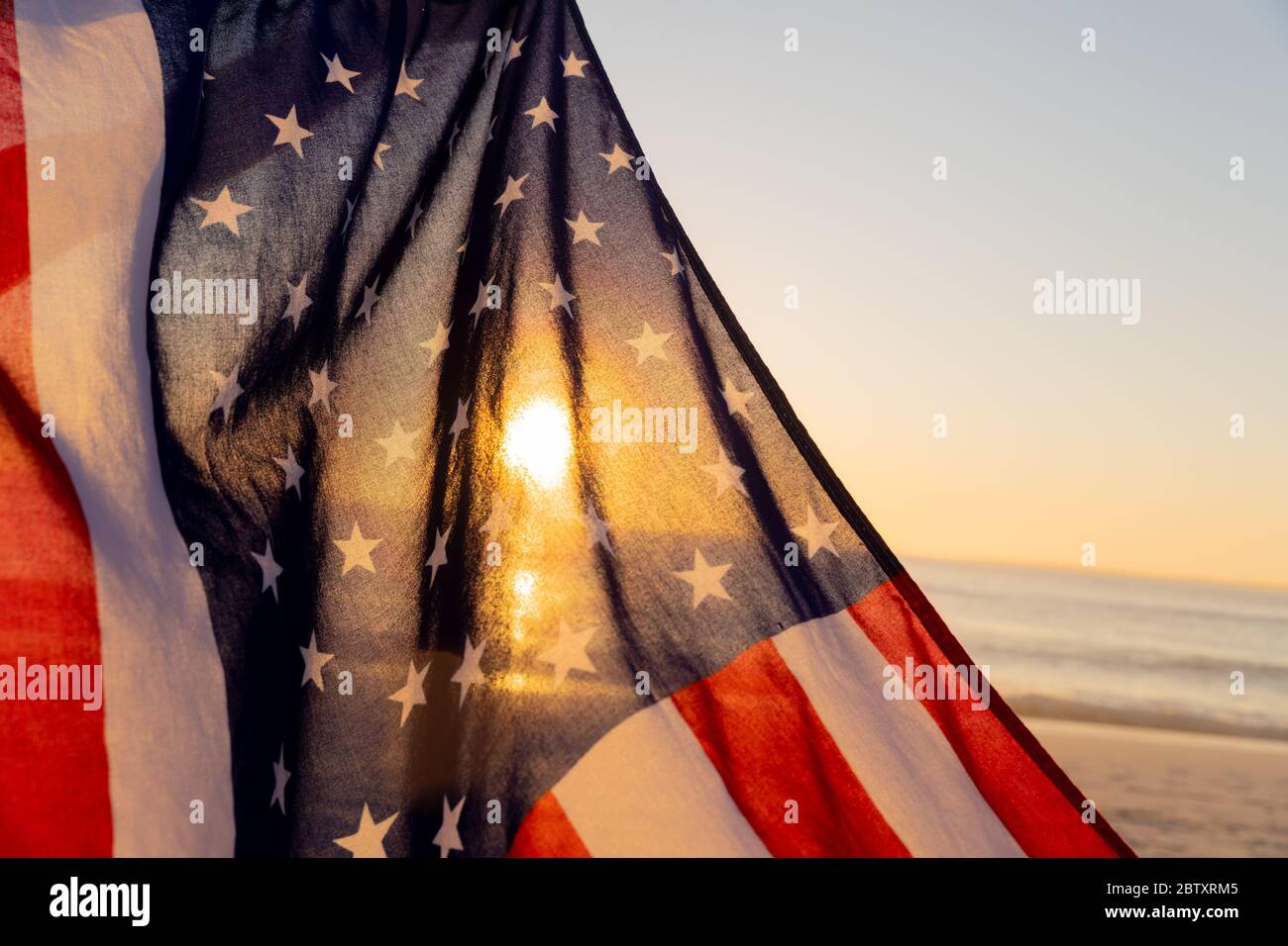The sun seen through an US flag on a beach, during a sunset with yellow ...