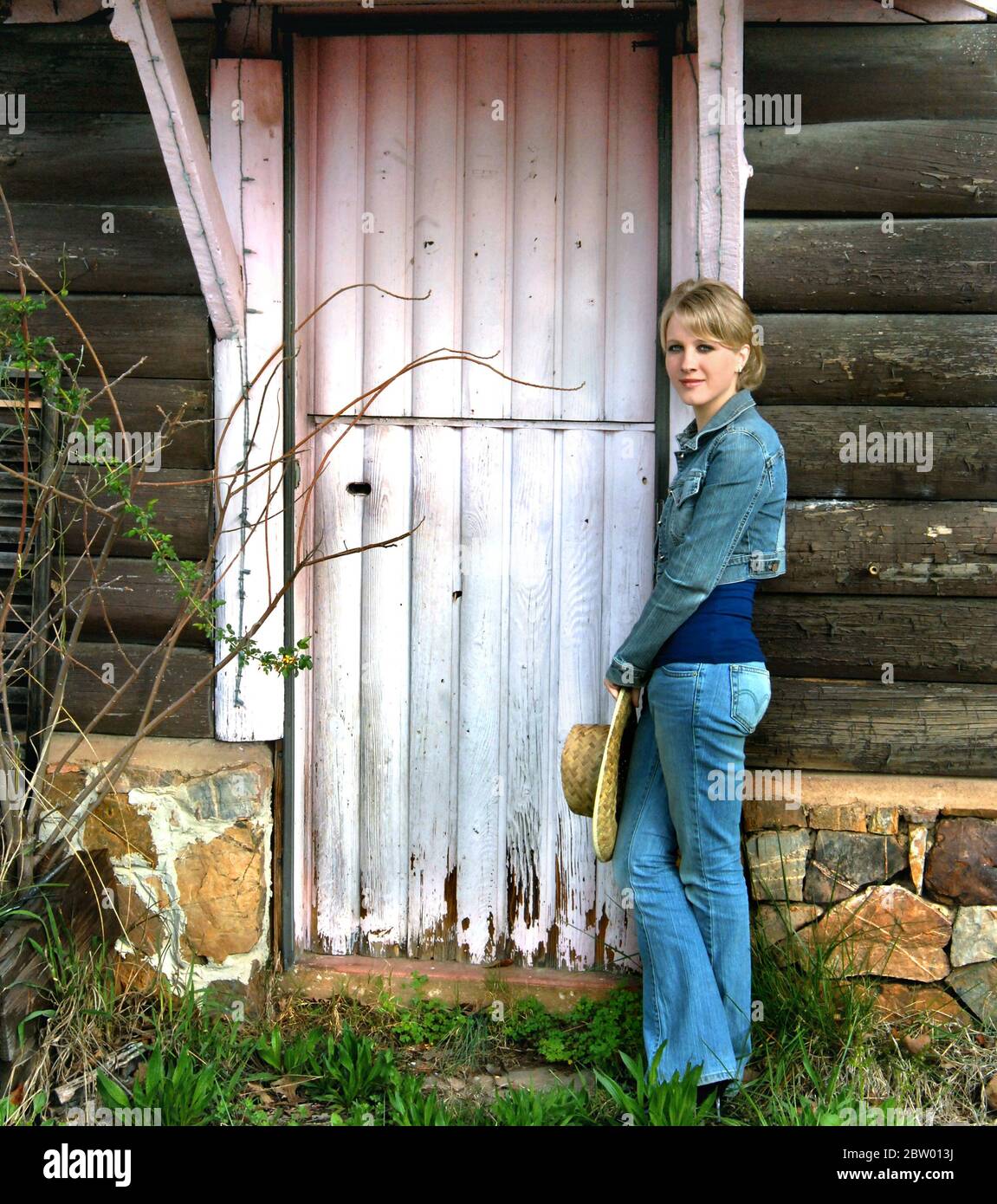 Young woman leans against a rustic log house that has a pink door. She ...
