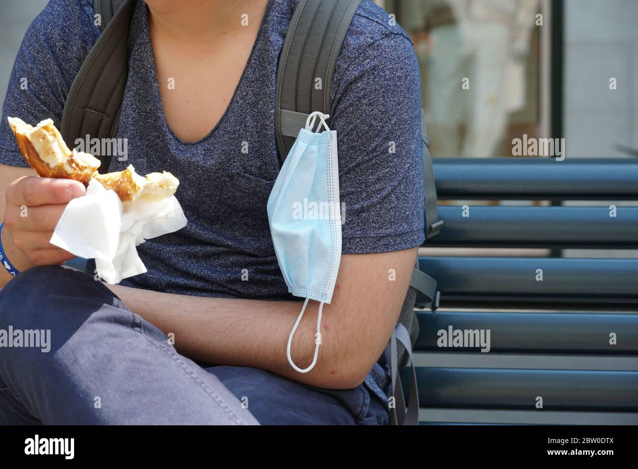 a teenager eating a breztel, his N95 face mask attached to his backpack ...