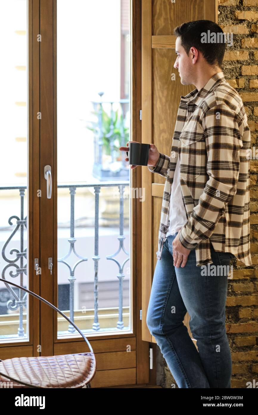 Young man chilling at home looking through the window to the street ...