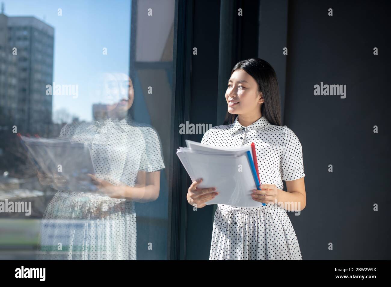 Young brunette female holding papers, looking into the window, smiling ...
