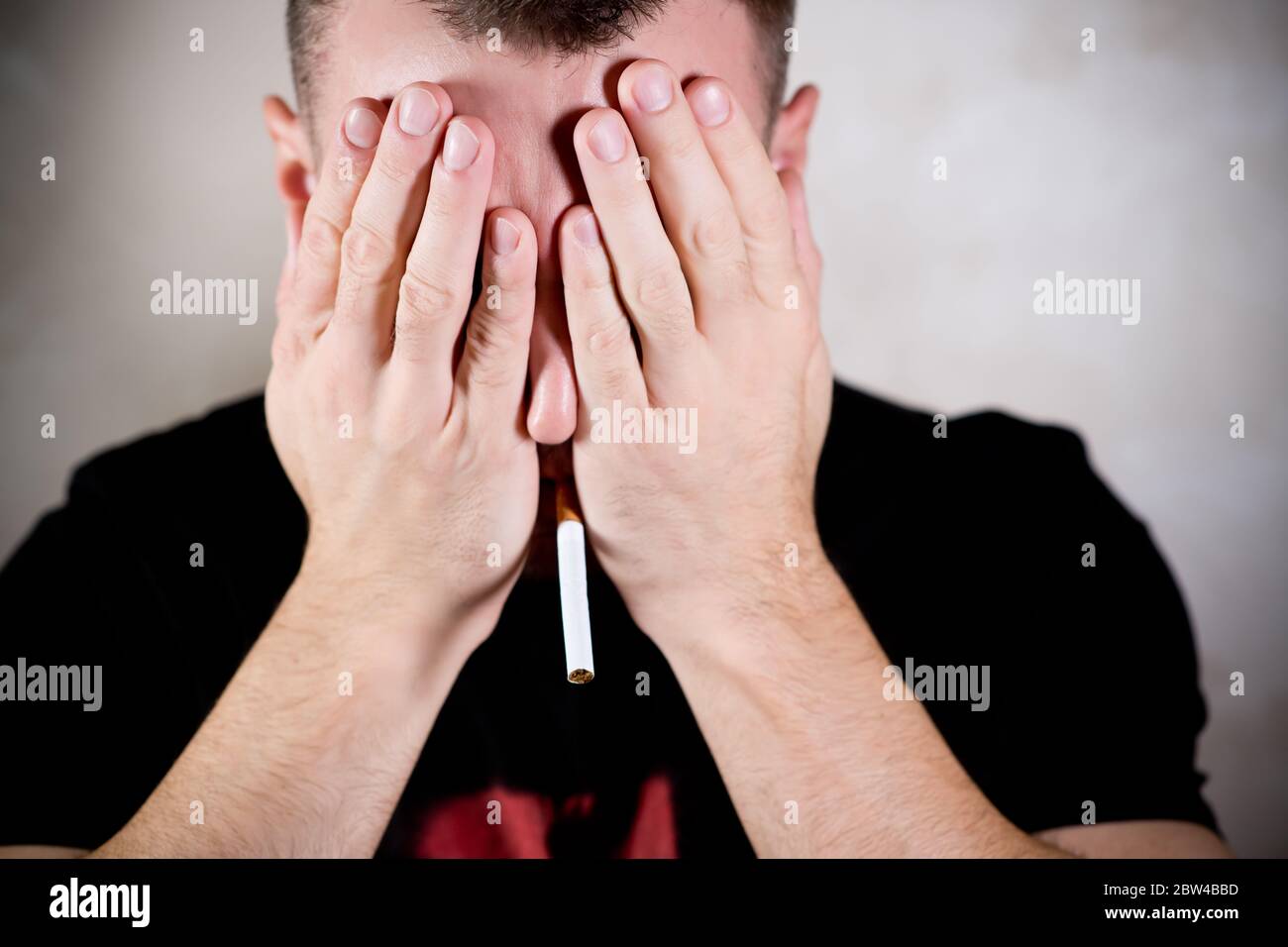 man with a cigarette in his mouth shyly covers his face with his palms ...