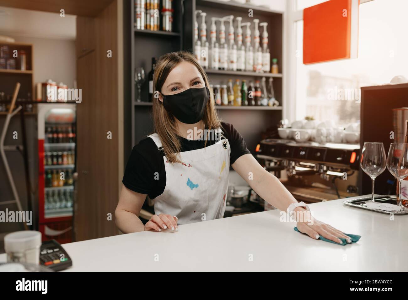 A female barista in a medical black face mask smiles and cleaning the ...