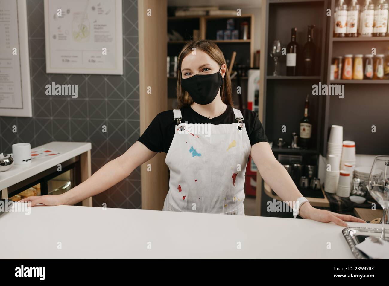A happy female barista in a medical black face mask smiles and waiting ...
