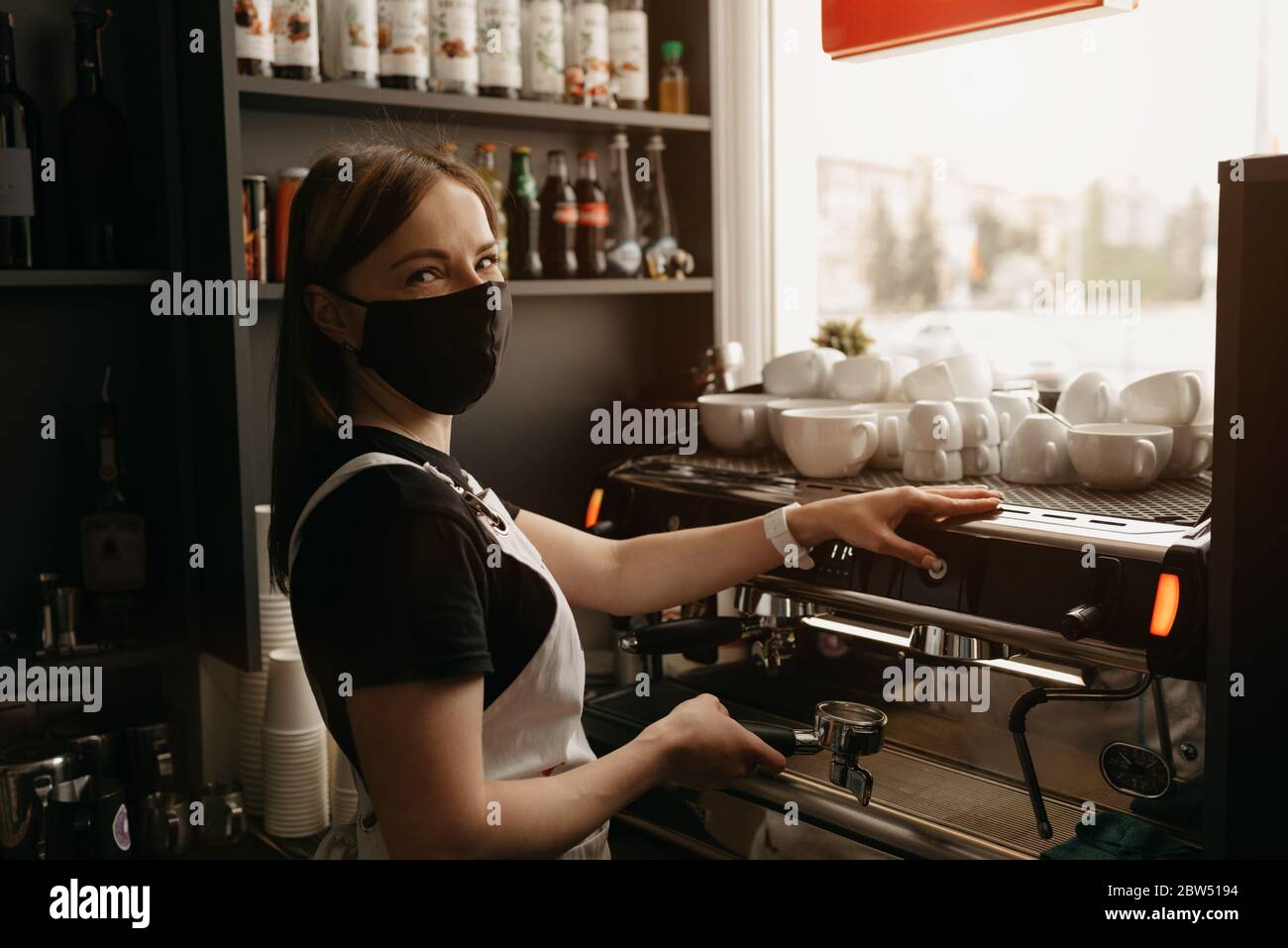 A happy female barista in a medical black face mask makes a cup of ...