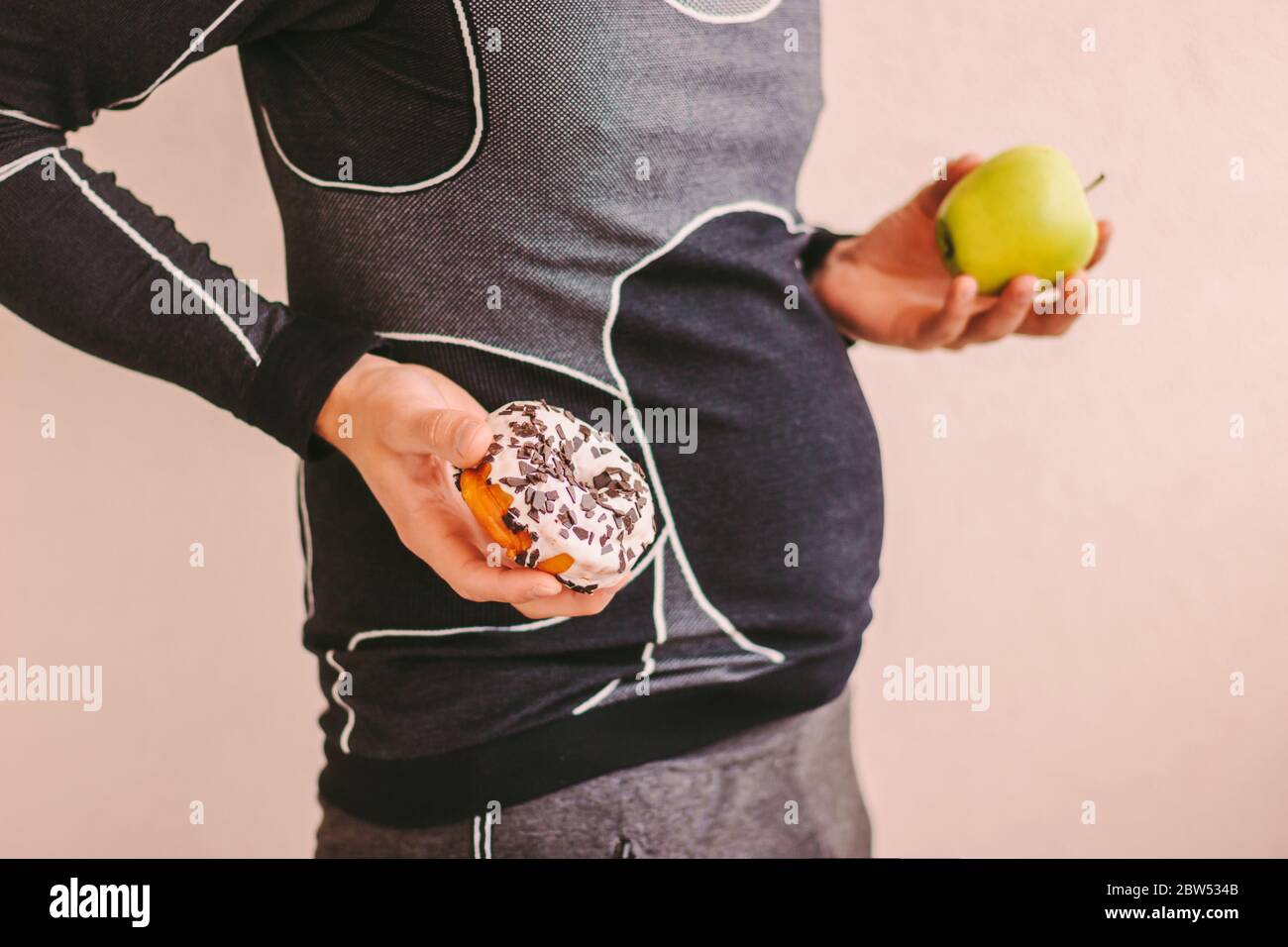 Closeup fat young sports man holding sweet donut and fresh apple in ...