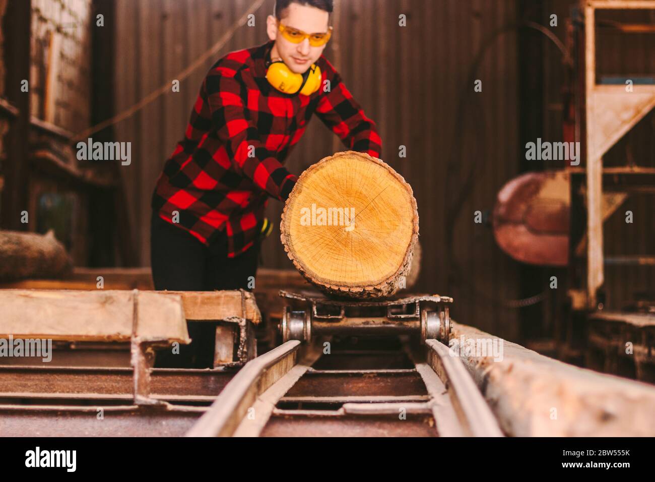 Young workman in protective glasses, headphones moving cut tree log to ...