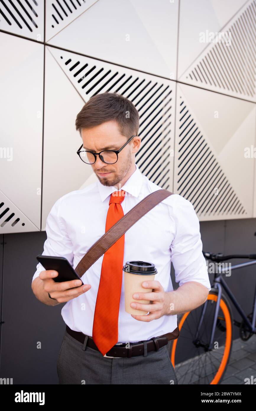 Young serious businessman scrolling in smartphone and having coffee ...