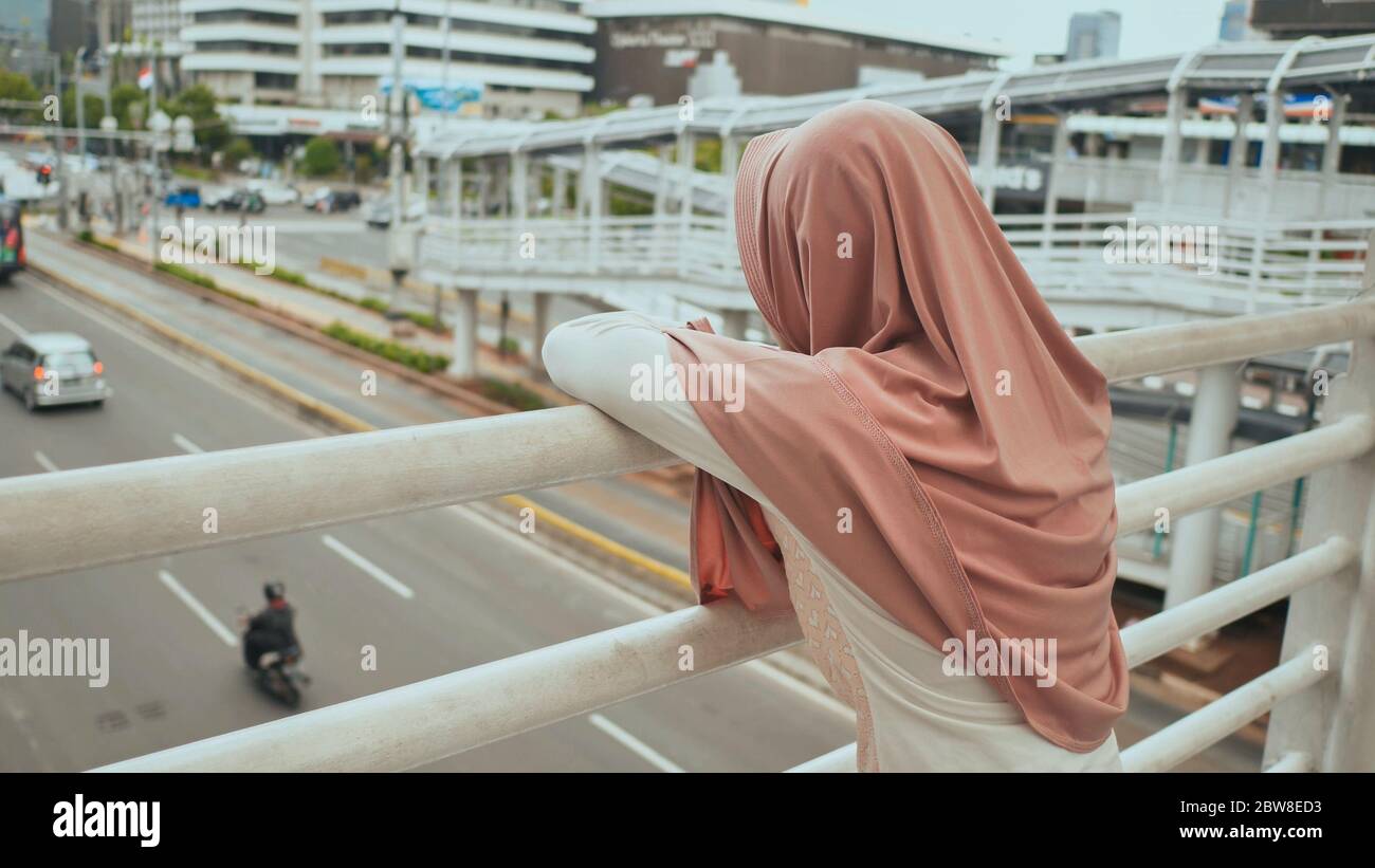 A young, sad Muslim girl stands on a bridge over road traffic in ...