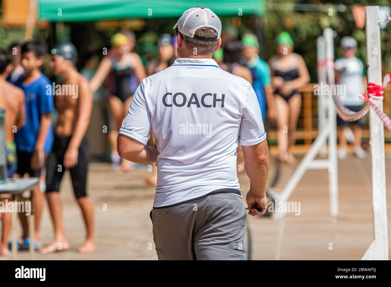 Back Of Male Swimming Coach Walking On The Poolside Toward His Young
